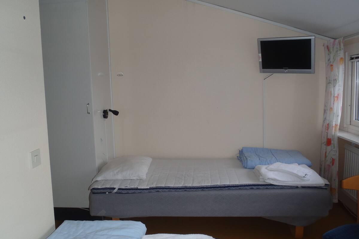 Quarto pequeno com uma cama de solteiro, televisão fixada na parede, porta e janela com cortinas.