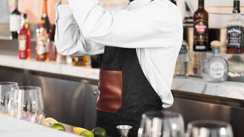 Um barman preparando bebidas num bar, com uma variedade de garrafas e copos ao fundo.