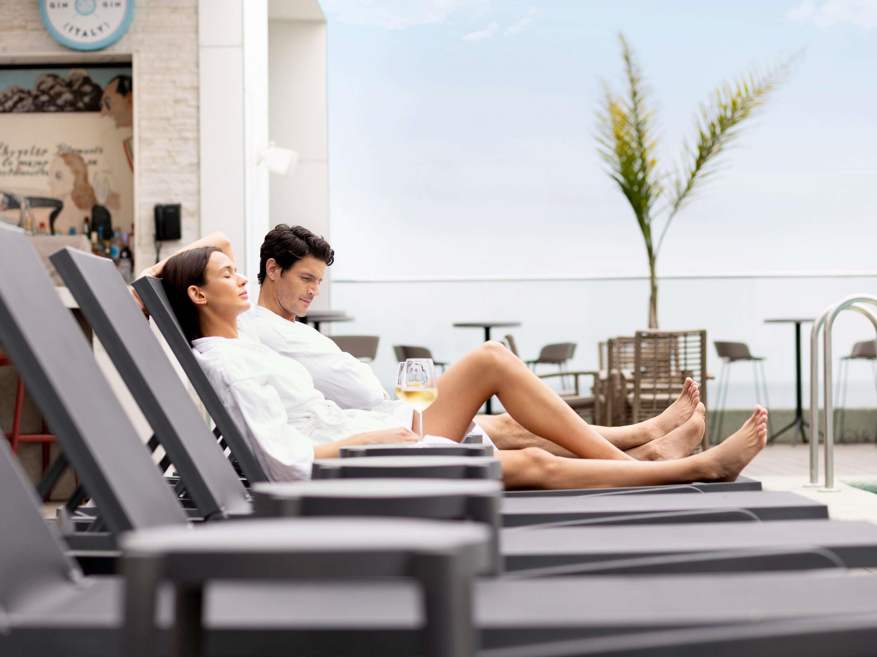 Um casal relaxando em espreguiçadeiras, com uma taça de vinho, apreciando a vista de uma área de piscina com cadeiras e uma palmeira.