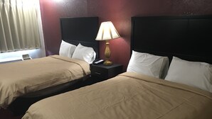 Quarto de hotel com duas camas, cada uma com colcha bege e almofadas brancas. Há uma mesa de cabeceira com abajur entre as camas.
