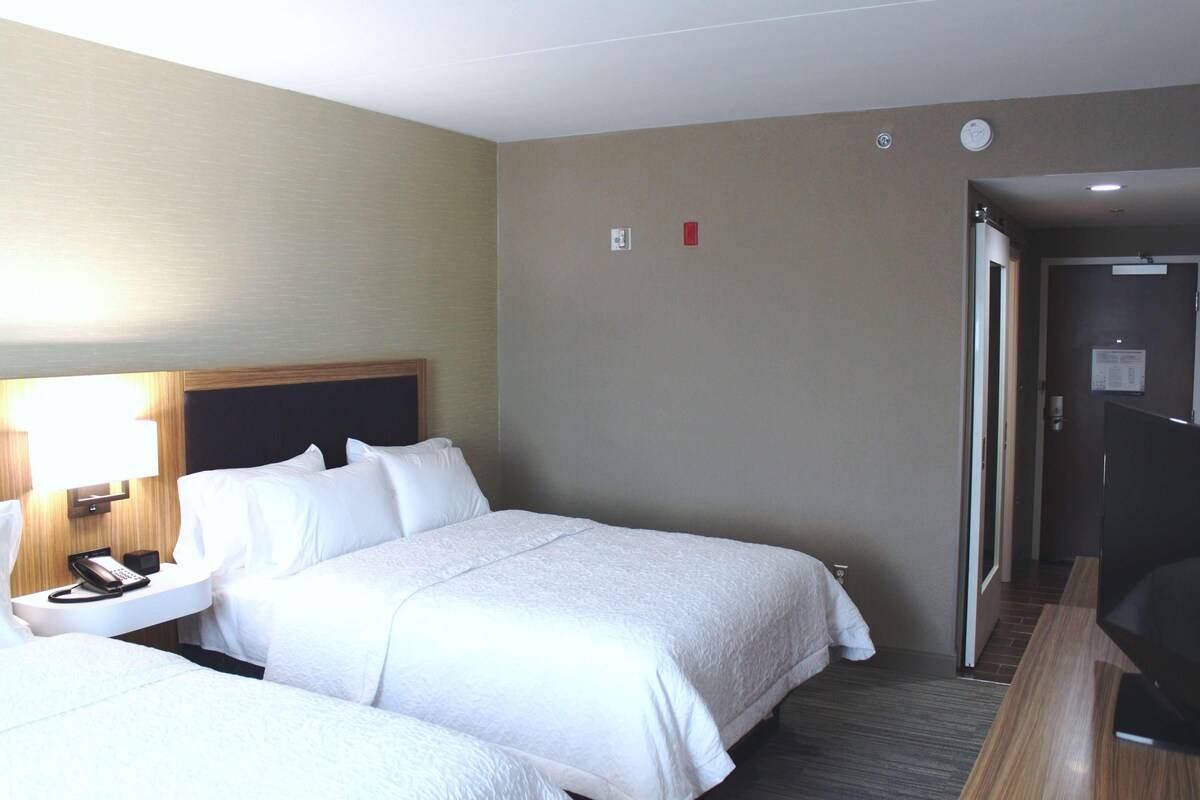 Quarto de hotel com duas camas, uma mesa de trabalho com luminária, televisão e relógio na parede.