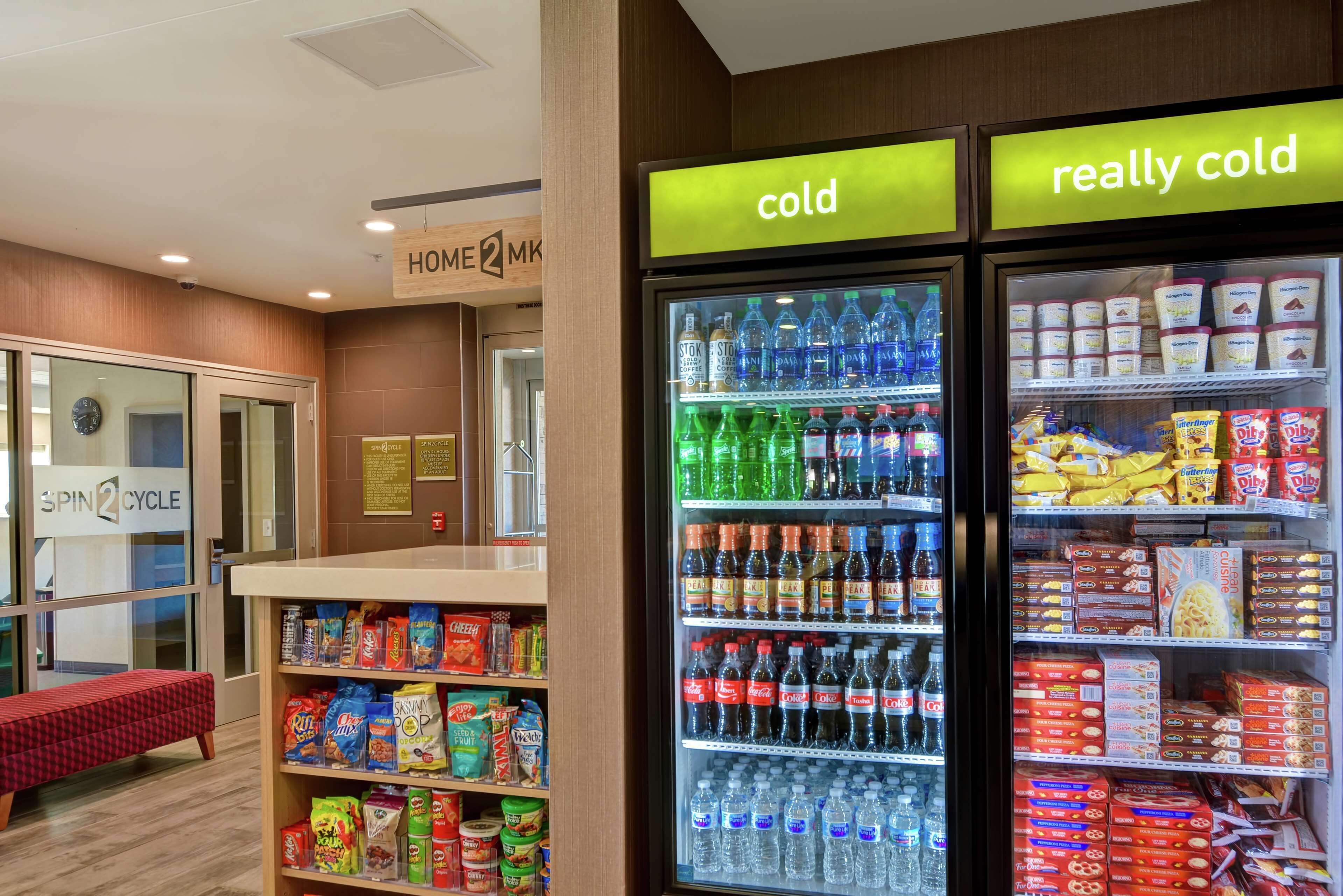 Um saguão de hotel com balcão de recepção, um banco e duas geladeiras identificadas como 'frio' e 'muito frio', repletas de bebidas e lanches.