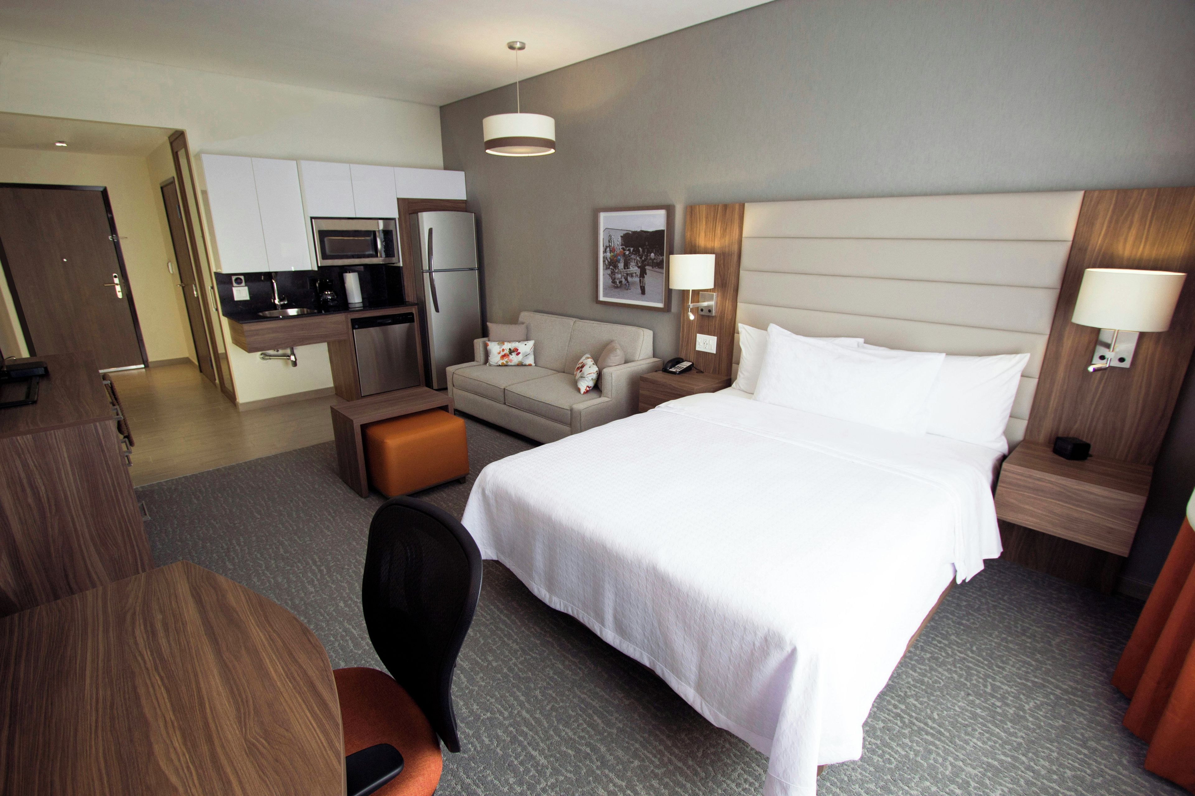 Quarto de hotel com cama, escrivaninha, cadeira, sofá, mesa de cabeceira, luminária, televisão na parede, micro-ondas, geladeira e porta para o banheiro.