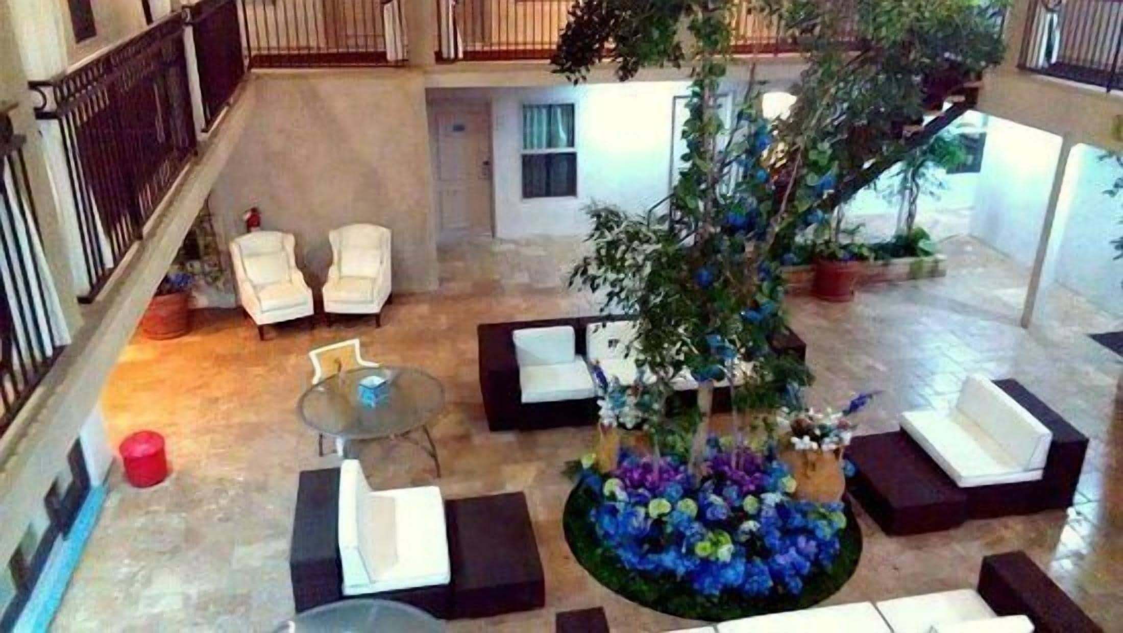 Amplo espaço interno com uma árvore central rodeada por flores azuis e roxas, sofás brancos e uma mesa de vidro.