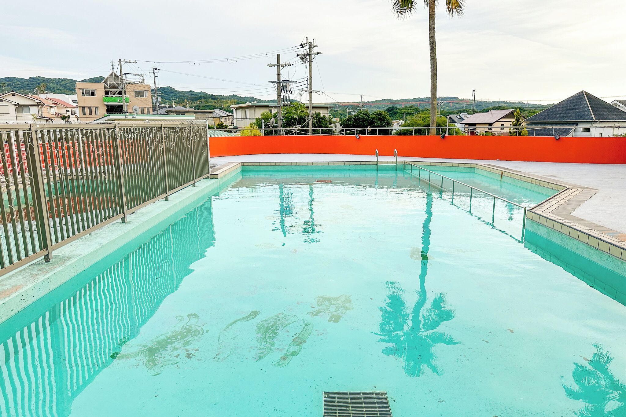Uma piscina com superfície de água azul cristalina, cercada por um muro vermelho e uma grade de metal.