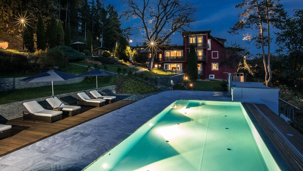 Piscina com espreguiçadeiras e deck de madeira, cercada por árvores e um prédio com várias varandas.