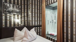 Quarto de hotel com cama, almofadas, uma imagem emoldurada da paisagem urbana, papel de parede listrado e área de chuveiro com divisória de vidro.