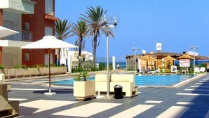 Uma área de piscina com espreguiçadeiras e guarda-sóis, um edifício com varandas e uma vista ampla do mar.