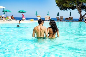 Um casal está em uma piscina, com cadeiras de espreguiçadeira e guarda-sóis ao fundo.