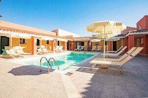 Uma área de piscina com espreguiçadeiras e guarda-sóis, cercada por edifícios com telhados de azulejos.