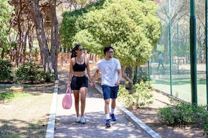 Duas pessoas caminham por um caminho pavimentado em um parque, com árvores e uma cerca ao fundo.