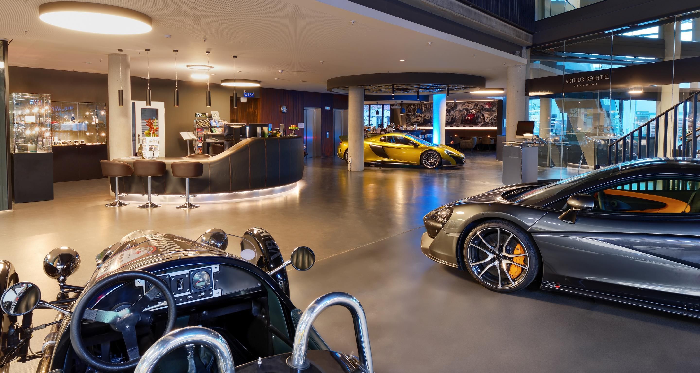 Interior moderno com balcão de recepção, área de estar e exposição de carros de luxo.