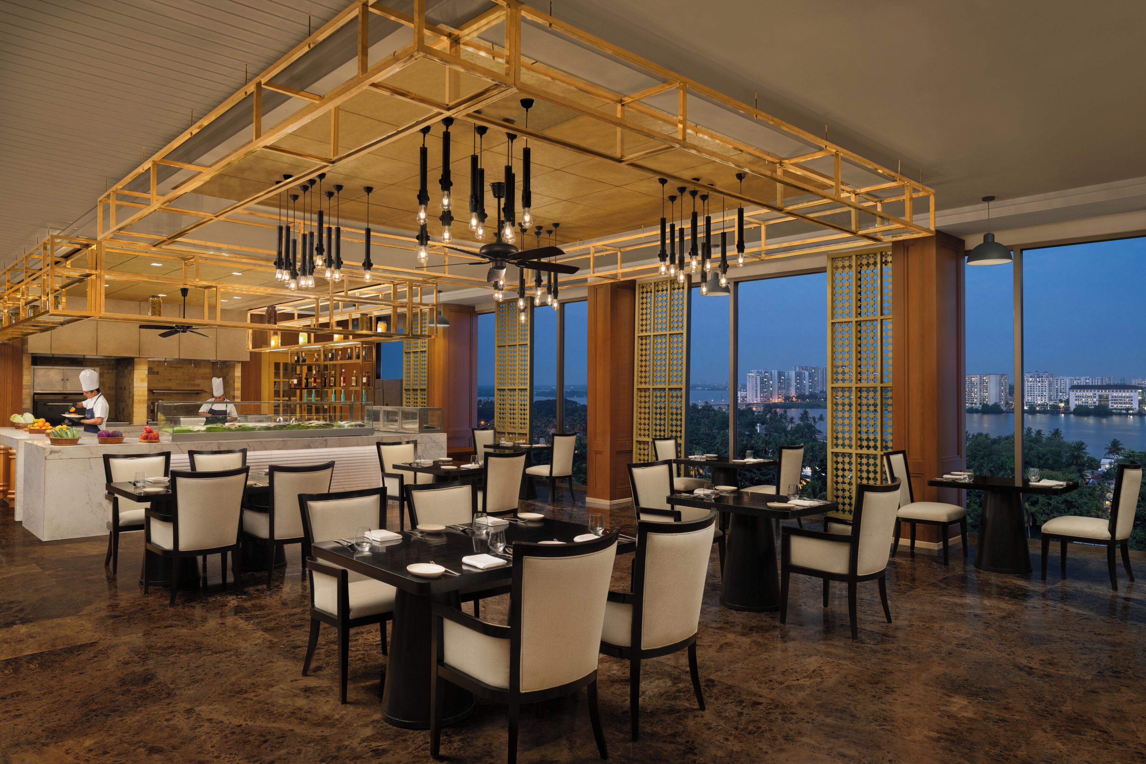 Um restaurante com várias mesas e cadeiras, uma área de cozinha e grandes janelas com vista para a cidade.