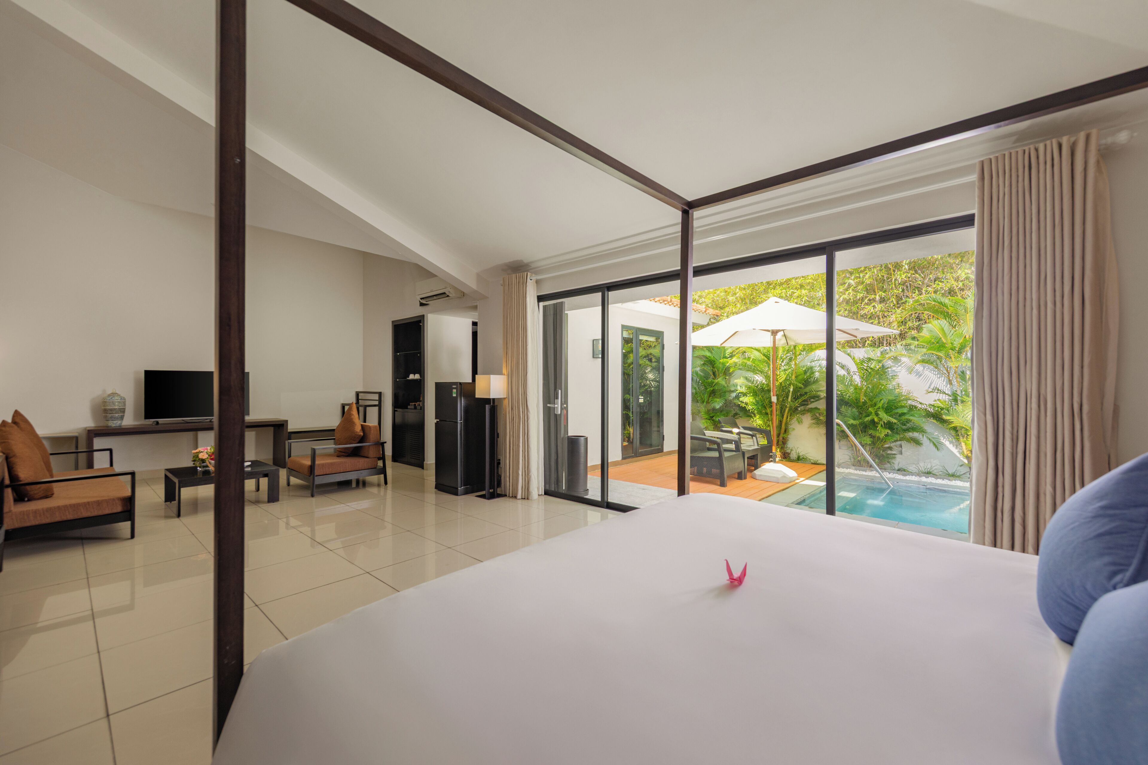 Quarto de hotel moderno com uma cama grande, televisão, área de refeições e vista para uma piscina ao ar livre.