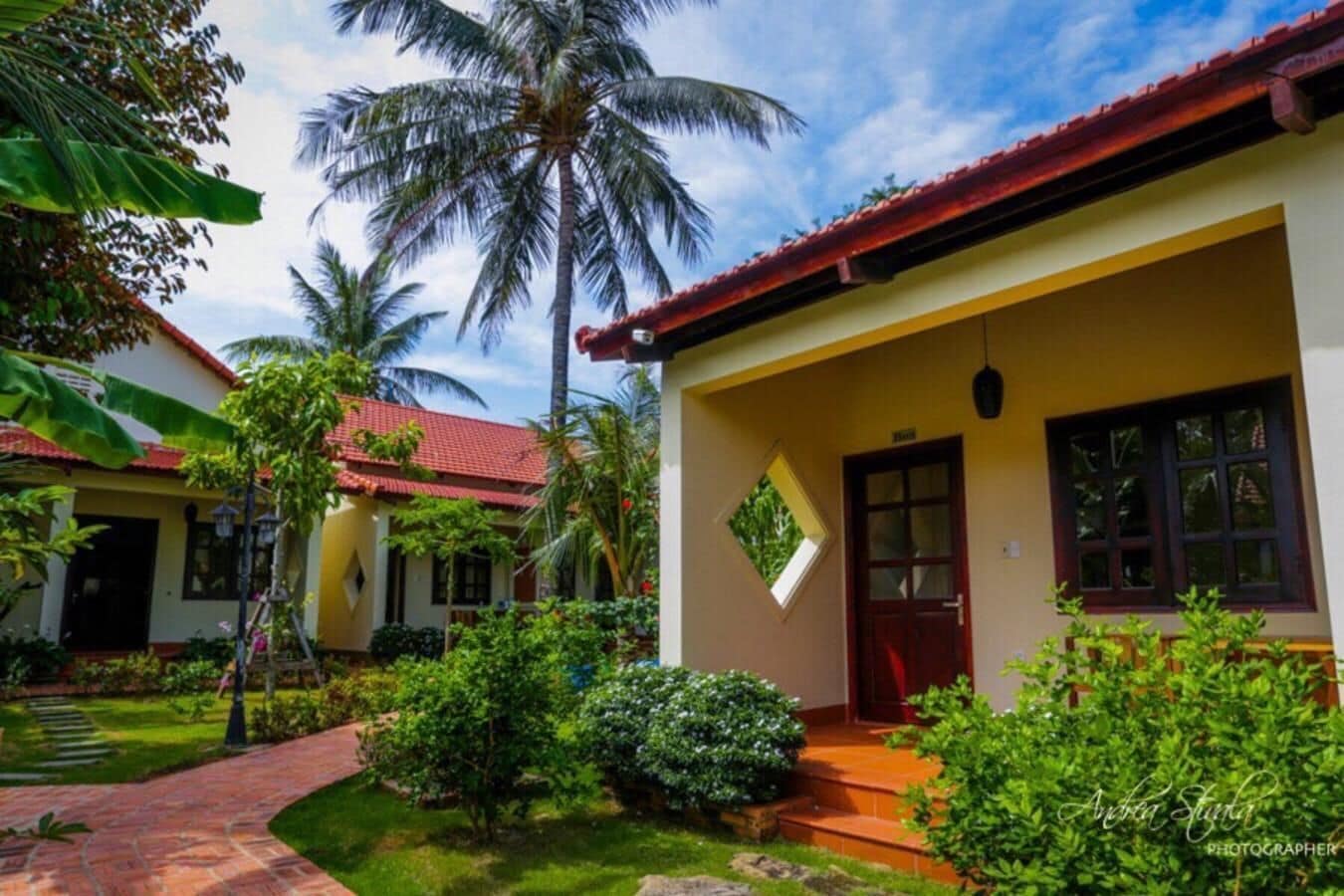 Um edifício de dois andares com telhado de telhas vermelhas, um pequeno alpendre e um jardim com palmeiras e arbustos.