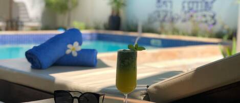 Área de piscina com uma espreguiçadeira, toalha enrolada, um copo de bebida amarela e óculos de sol em uma mesa pequena.