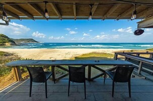 Uma mesa de madeira com cadeiras sob uma cobertura com luzes penduradas, com vista para uma praia e uma visão clara do oceano e colinas ao longe.