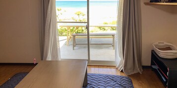 Quarto com cama de madeira, vista para a praia através de uma porta deslizante, uma mesa pequena e um cesto numa prateleira.