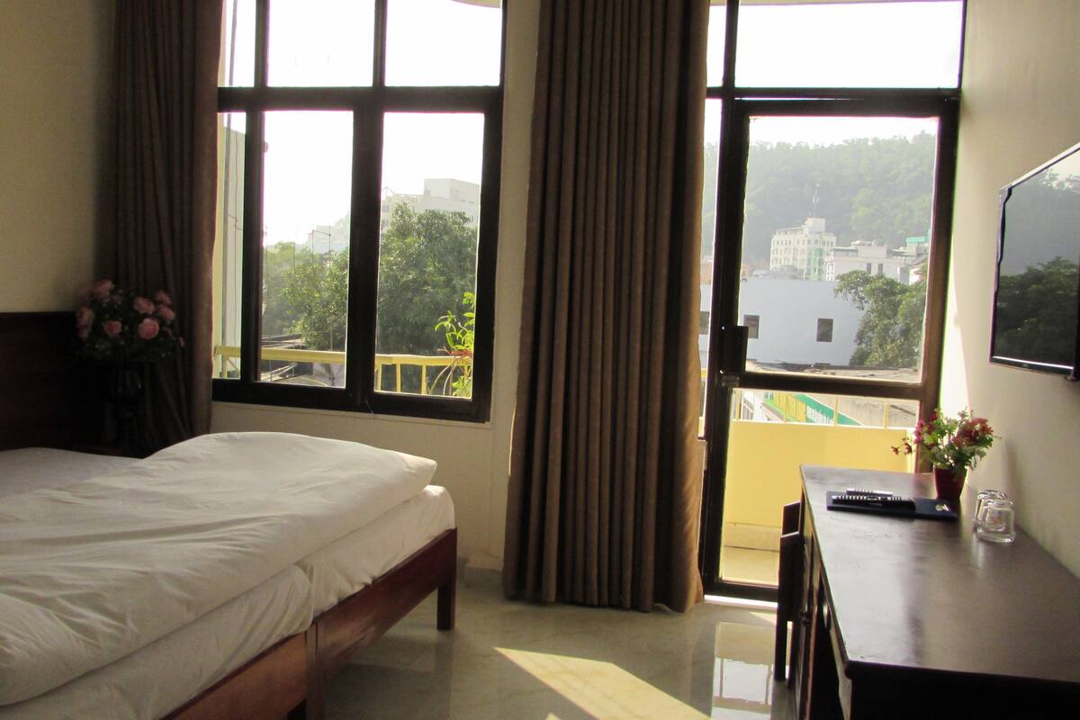 Quarto de hotel com cama, escrivaninha, cadeira e uma janela grande com cortinas.