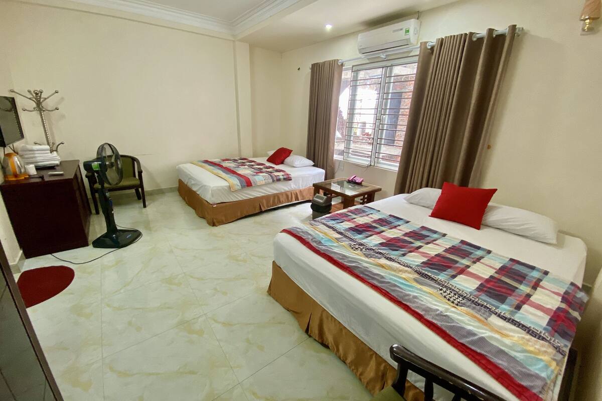 Quarto de hotel com duas camas, uma escrivaninha, uma cadeira, ar condicionado e uma janela com cortinas.