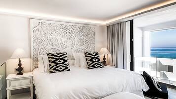 Quarto de hotel moderno com uma cama grande, duas luminárias de cabeceira, um painel decorativo na parede e vista para o oceano.