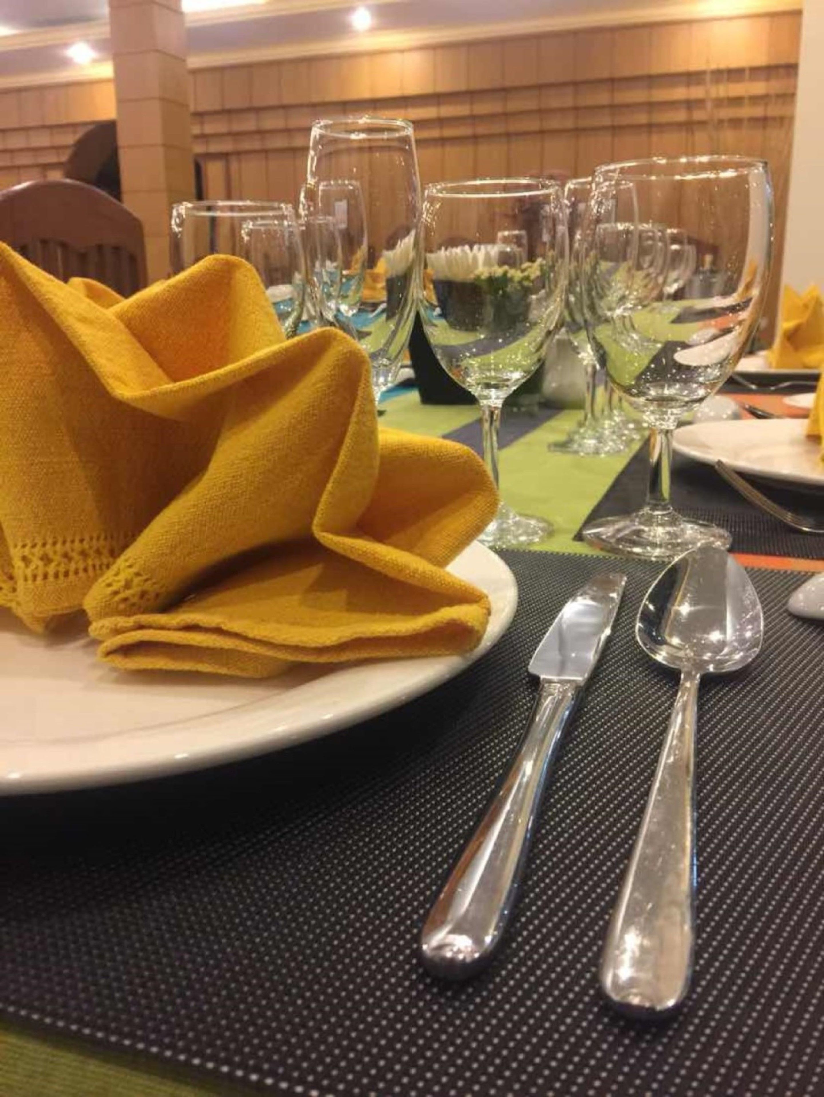 Uma mesa de jantar posta com uma toalha de papel amarela dobrada, um prato, talheres e copos de vinho.