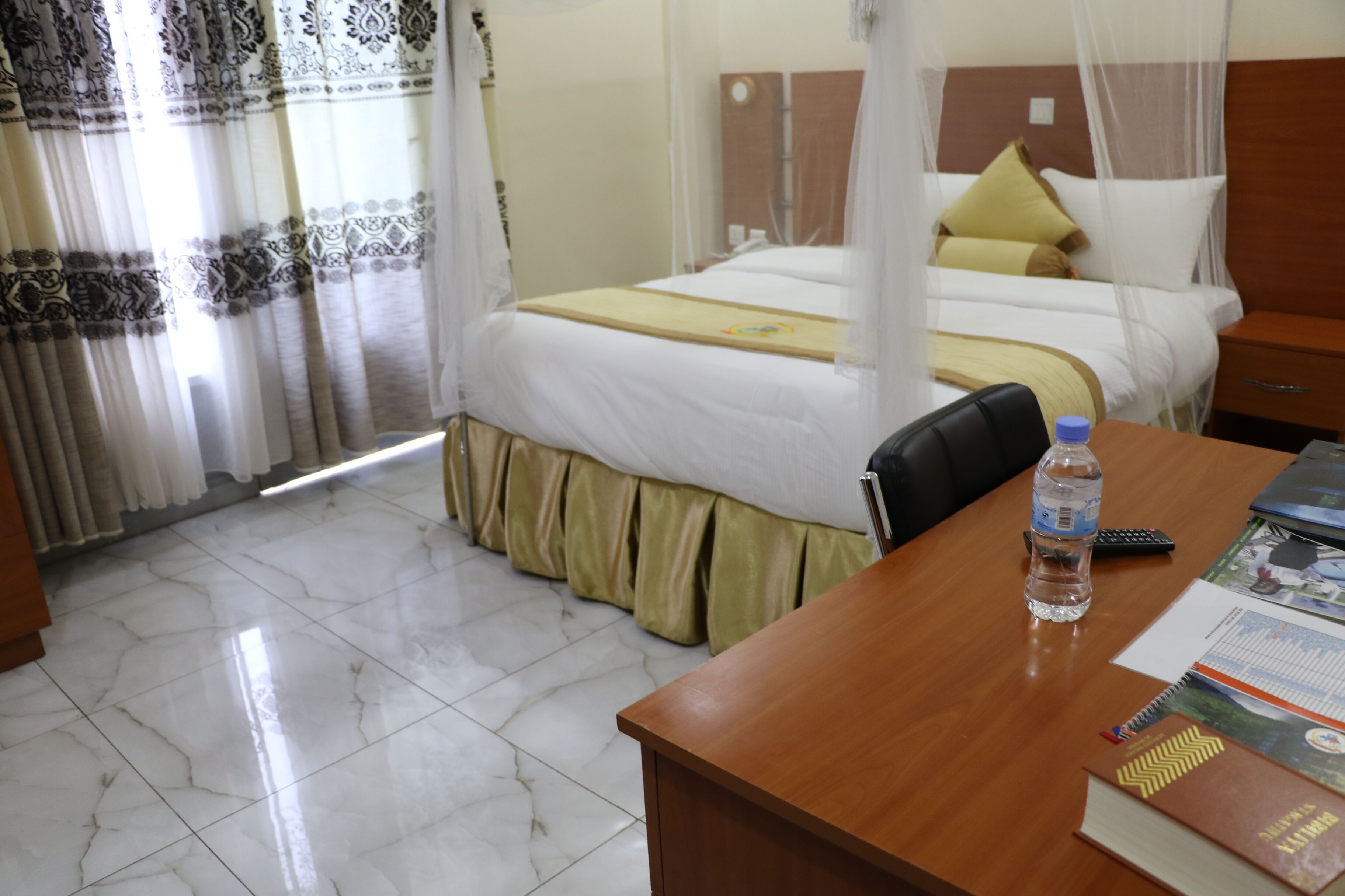 Quarto de hotel com cama, escrivaninha de madeira, cadeira, garrafa de água e caderno.