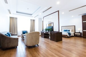 Quarto de hotel com piso de madeira, um sofá, uma cadeira, uma cama, uma televisão e uma escrivaninha.