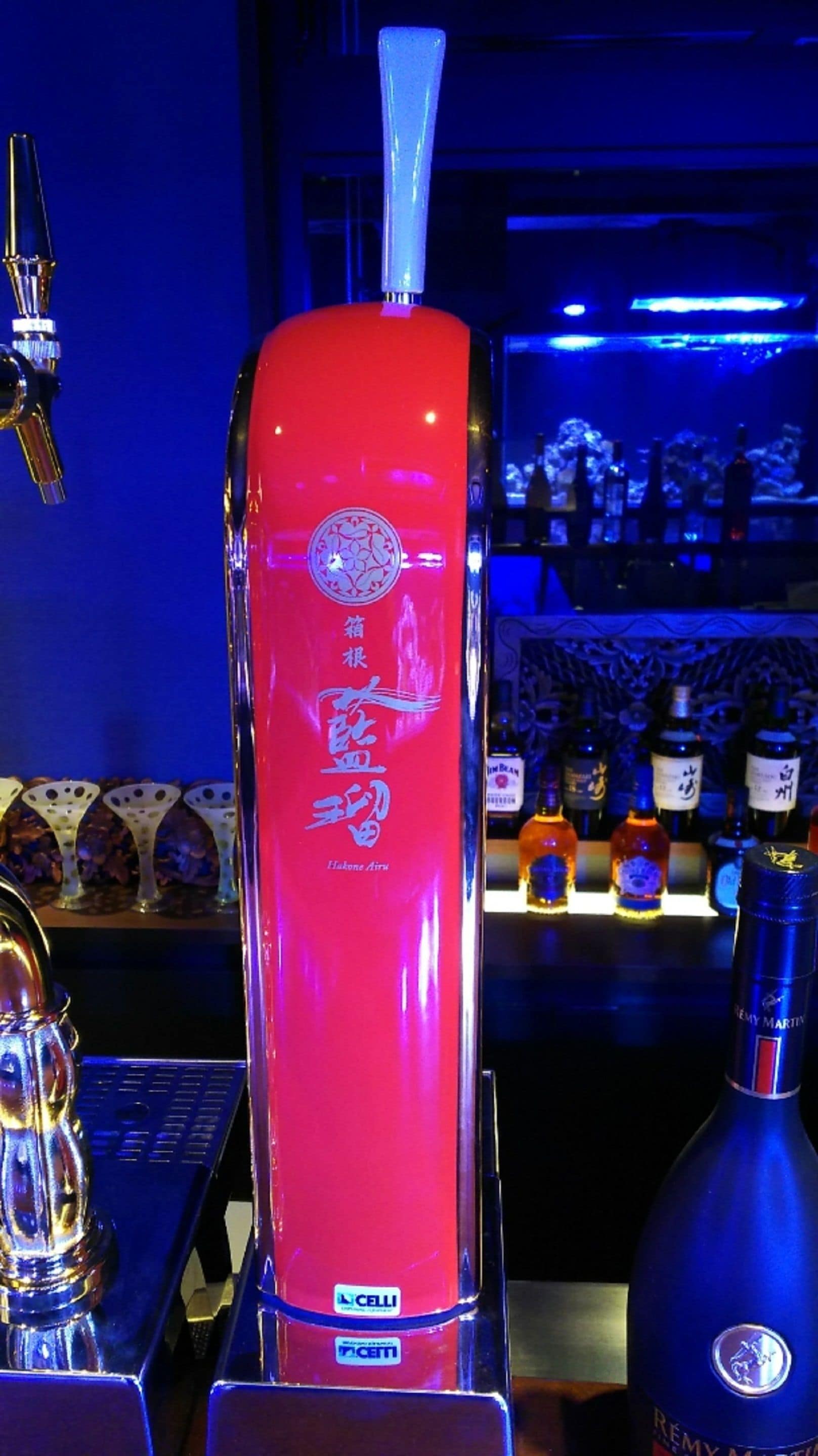 Uma torneira de bar vermelha com rótulo branco e uma garrafa de Remy Martin ao fundo.
