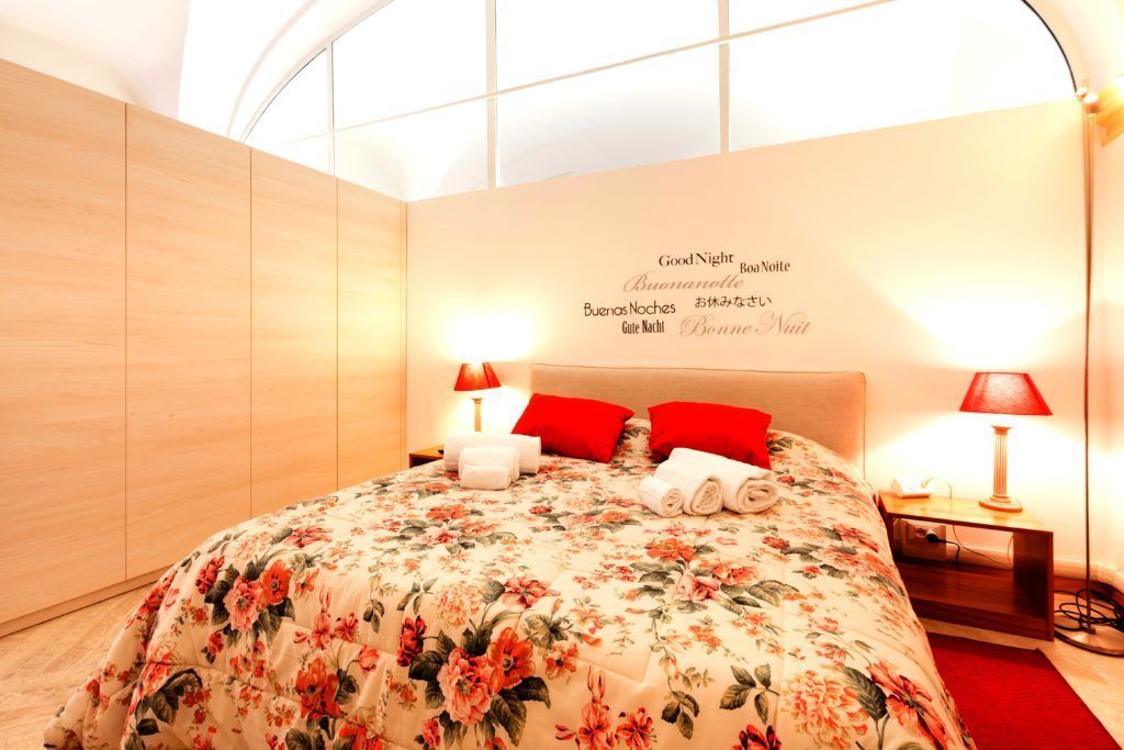 Quarto de hotel com colcha estampada com flores, almofadas vermelhas e parede com a inscrição 'Boa noite' em vários idiomas.