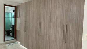 Quarto com um guarda-roupa espaçoso, banheiro visível através de uma porta aberta e piso ladrilhado.