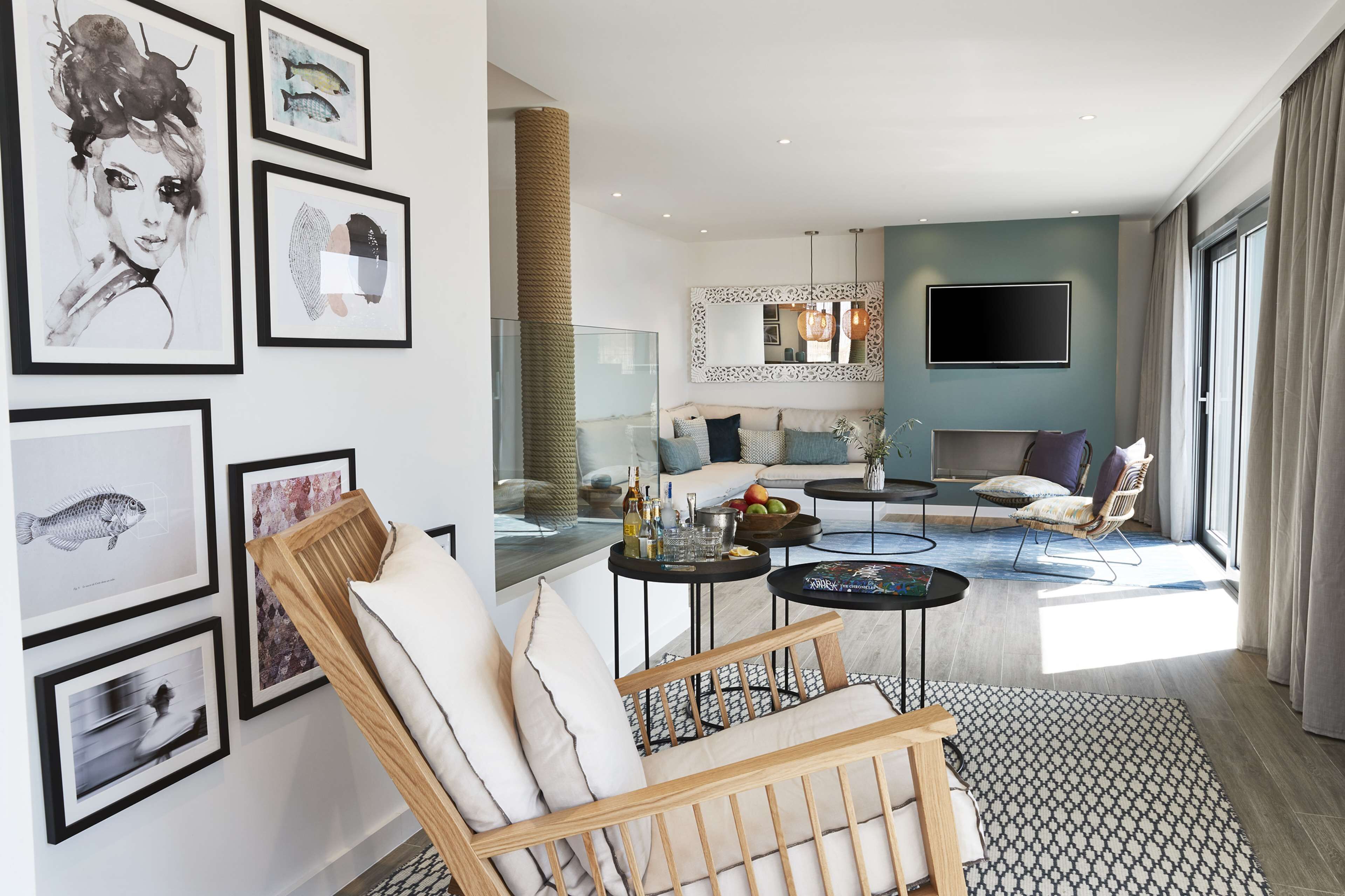 Uma sala de estar moderna com um sofá, poltronas, uma mesa de centro e uma televisão. Há quadros nas paredes e uma janela grande com cortinas.