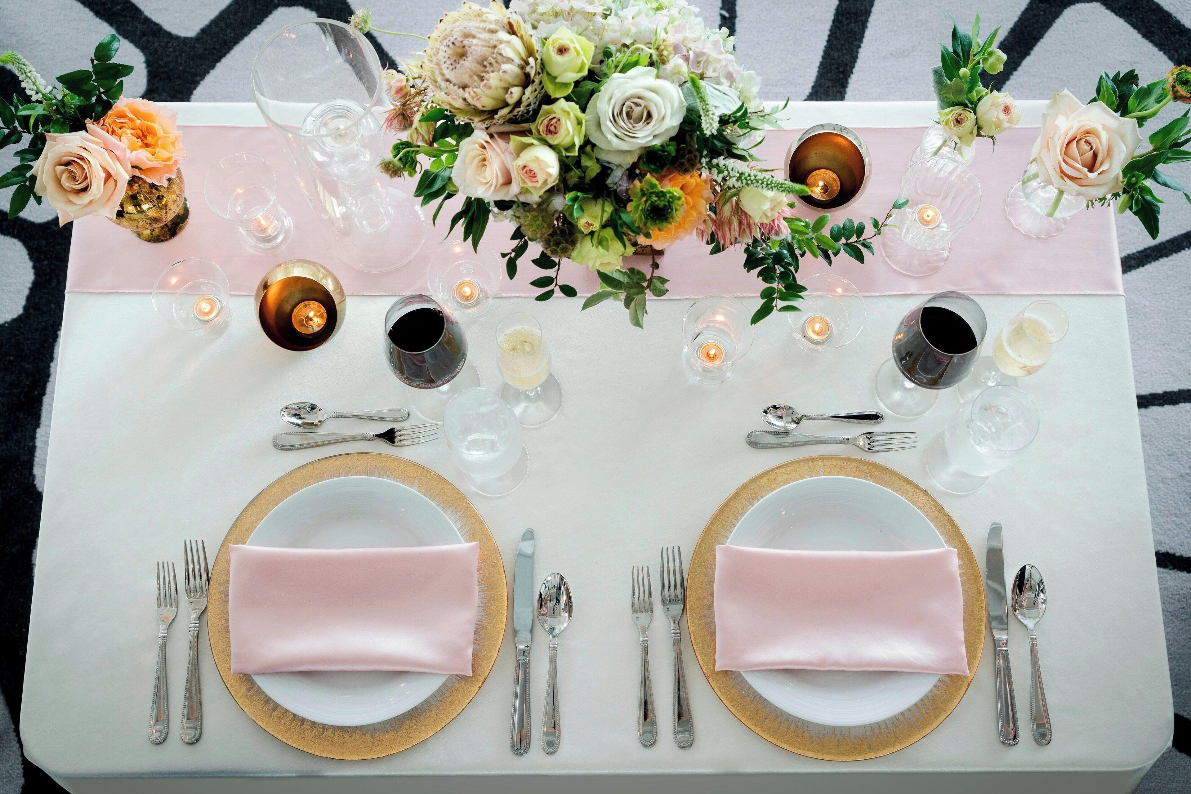 Mesa de jantar posta para dois, com pratos com detalhes dourados, guardanapos cor-de-rosa e talheres de prata.
