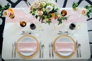 Mesa de jantar posta para dois, com pratos com detalhes dourados, guardanapos cor-de-rosa e talheres de prata.