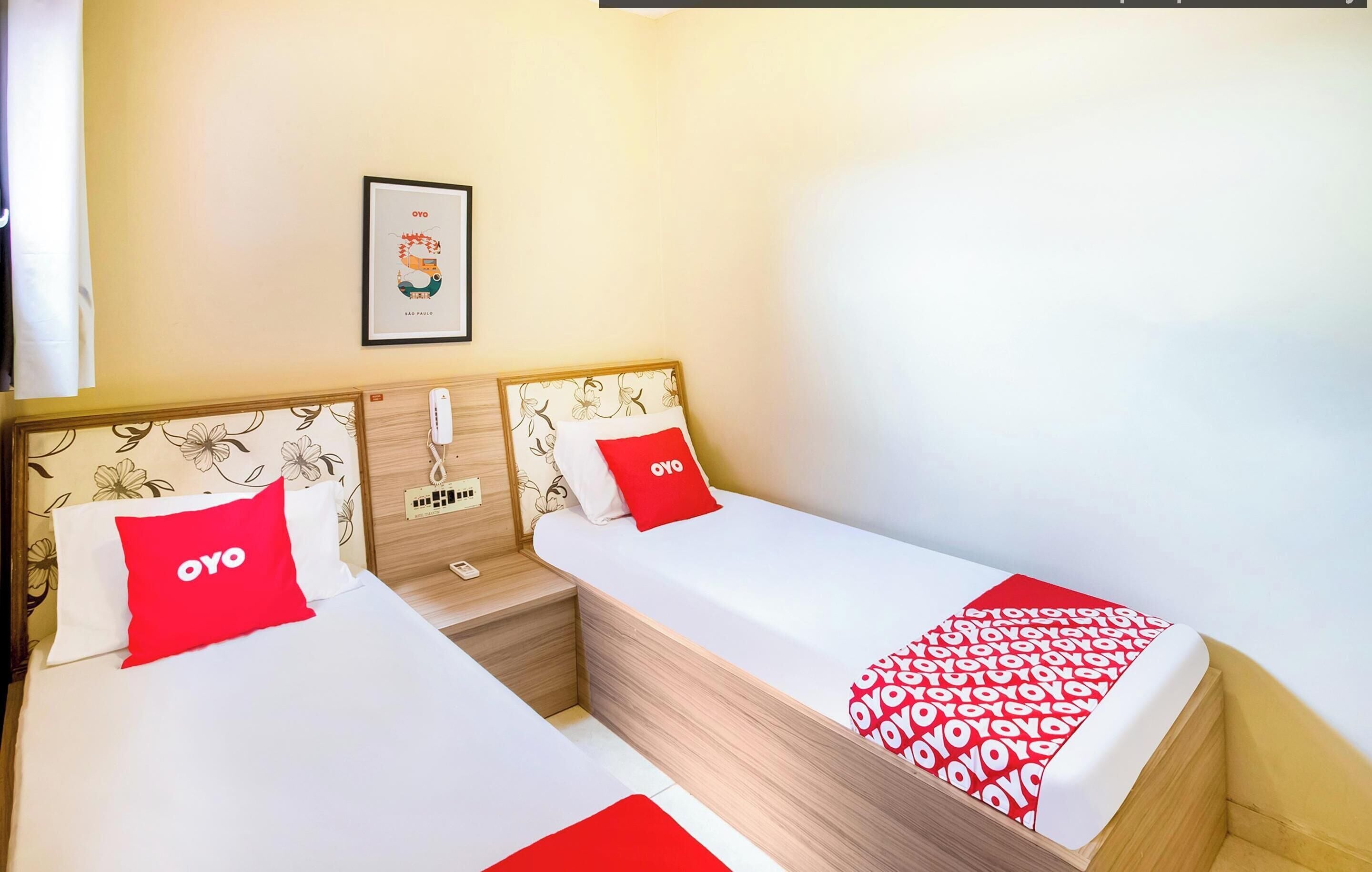 Quarto de hotel com duas camas, cada uma com travesseiro vermelho e colcha branca. Há um quadro na parede e uma mesa de cabeceira com telefone e tomada.