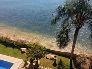 Visão ampla de uma praia com piscina, palmeiras e área de jardim com pedras e plantas em vasos.