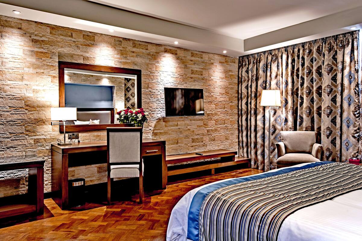 Quarto de hotel com parede de pedra, cama com colcha listrada, escrivaninha com cadeira, televisão e abajur.