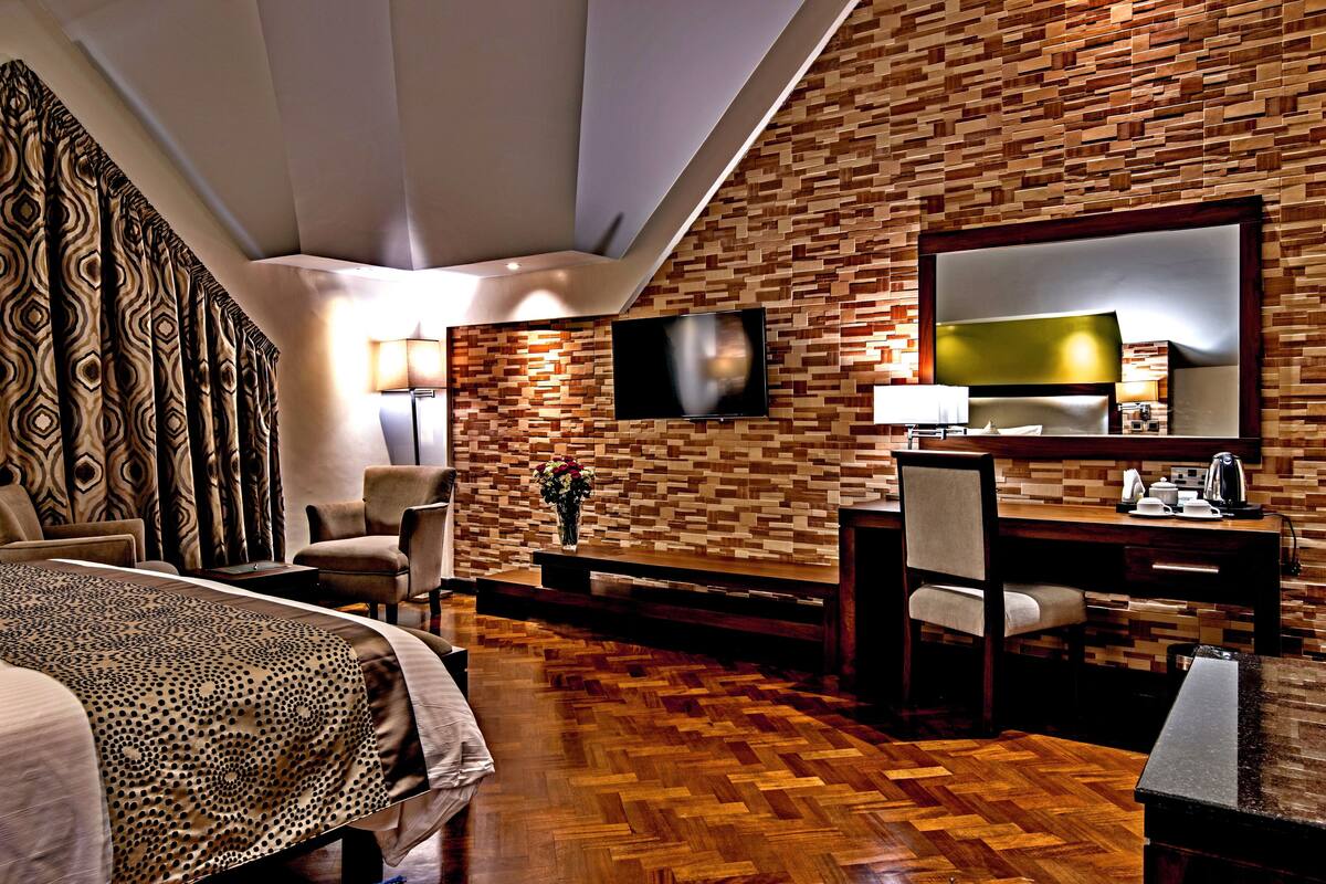 Quarto de hotel com design moderno, apresentando um chão de madeira em padrão herringbone, uma cama grande com colcha estampada, uma escrivaninha com cadeira, uma TV de tela plana na parede e um espelho grande sobre a escrivaninha.
