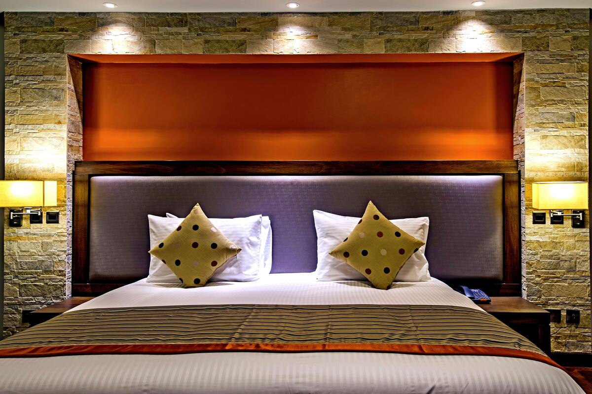 Quarto de hotel com parede de pedra, uma cama grande com roupa de cama branca e almofadas estampadas, e duas luminárias de cabeceira.