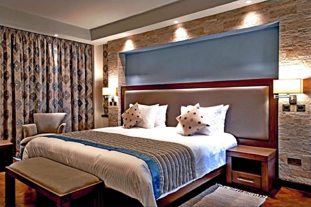 Quarto com cama grande, cabeceira, banqueta, cadeira e parede de pedra.