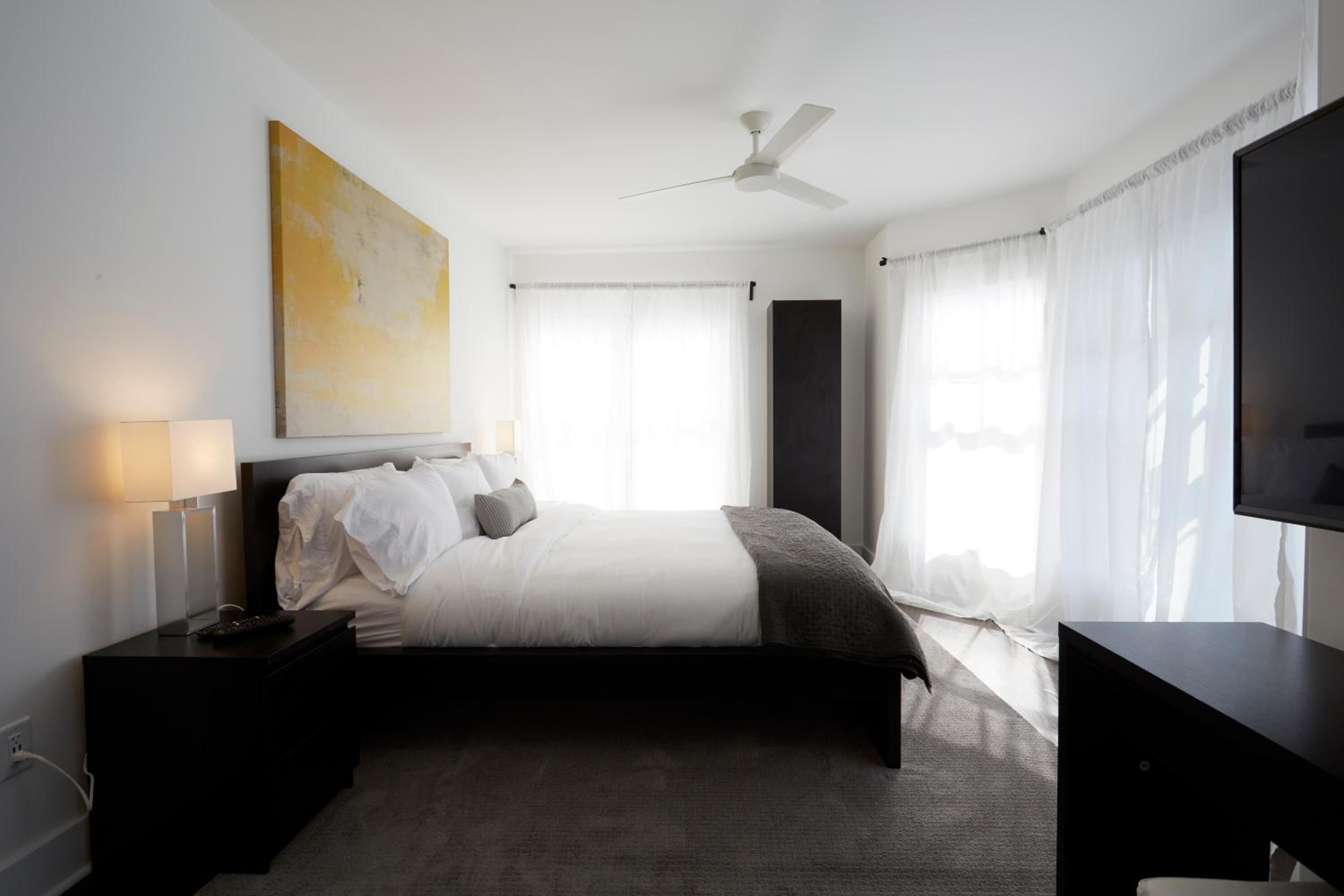 Quarto moderno com cama, mesas de cabeceira, ventilador de teto e uma janela grande com cortinas.