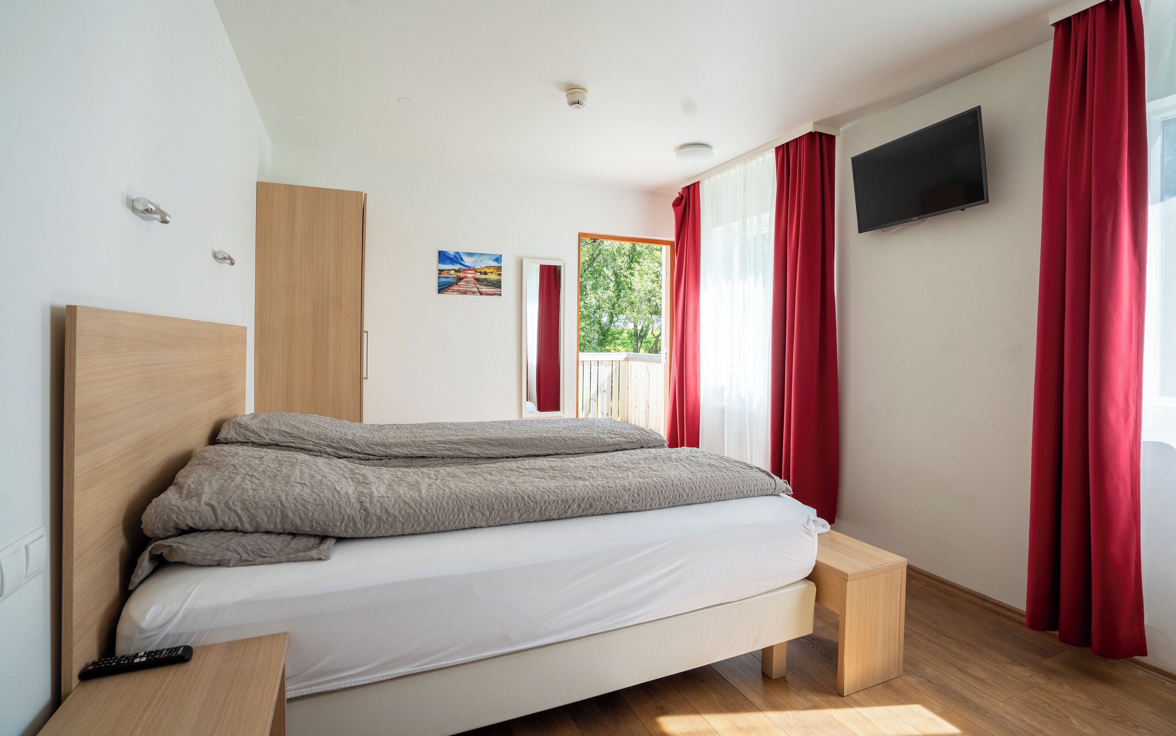 Quarto com cama, televisão fixada na parede e janela com cortinas vermelhas.