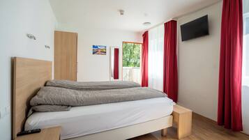 Quarto com cama, televisão fixada na parede e janela com cortinas vermelhas.