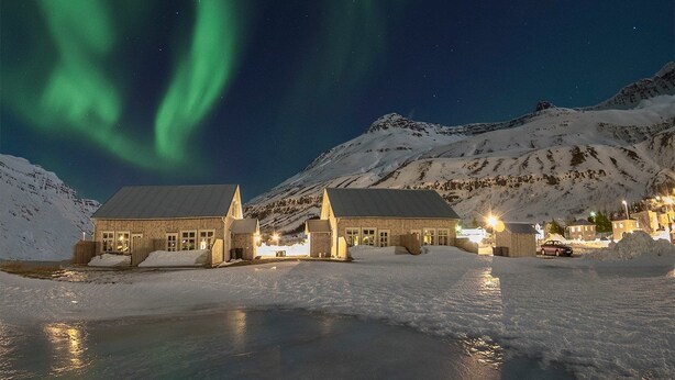 Um edifício coberto de neve com uma superfície aquática refletora, com montanhas nevadas ao fundo e a aurora boreal.