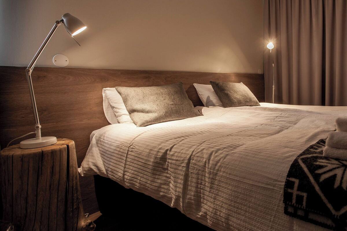 Cama bem arrumada com manta estampada, duas luminárias de cabeceira e uma mesinha de cabeceira de madeira.