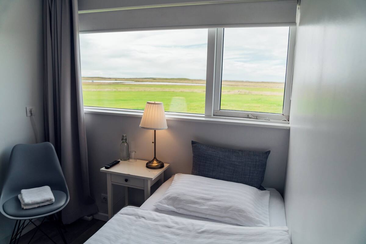 Quarto de hotel com uma cama, uma cadeira, uma mesinha de cabeceira, um abajur e uma janela com vista para um campo gramado.
