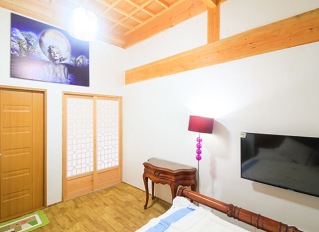 Quarto com cama, guarda-roupa de madeira, mesinha de cabeceira, televisão e luminária presa à parede.
