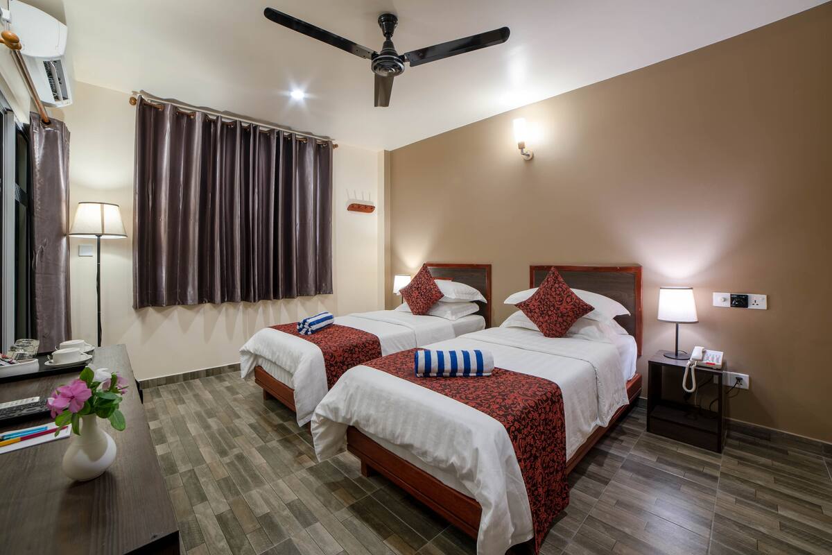 Quarto de hotel com duas camas, uma mesa com um vaso de flores, uma luminária e um ventilador de teto.