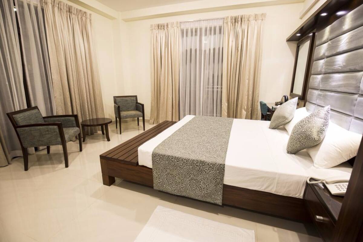 Quarto de hotel com uma cama grande, duas cadeiras, uma mesinha e um telefone ao lado da cama.