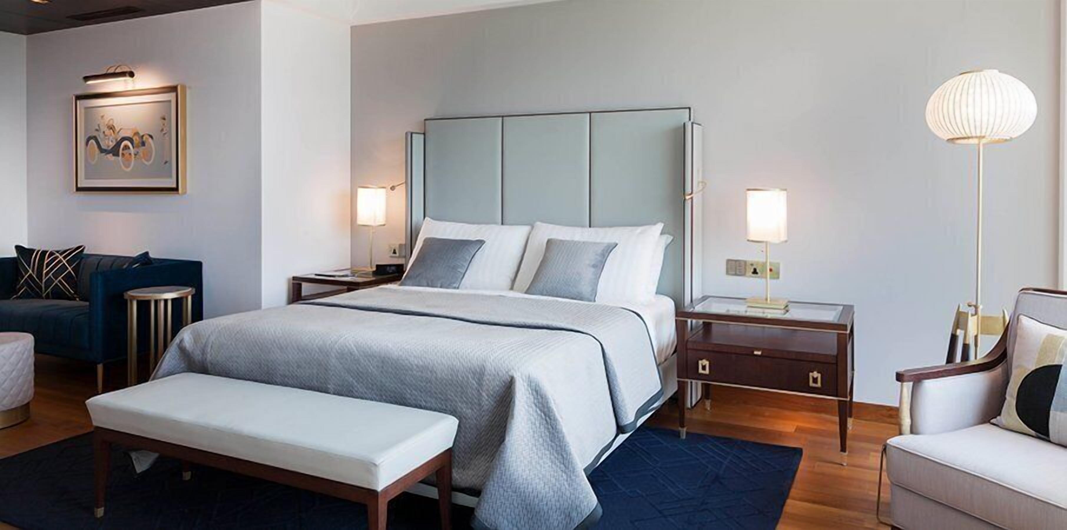 Quarto de hotel moderno com uma cama grande, mesas de cabeceira, um banquinho, um sofá e uma luminária.